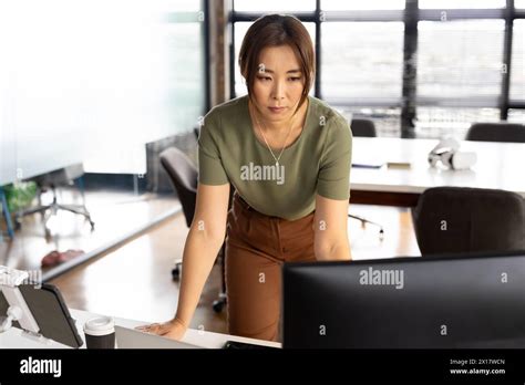 Middle Aged Asian Woman Standing Looking At Computer In A Modern Business Office Stock Photo