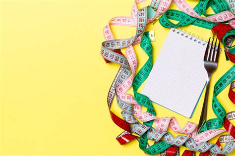 노란색 배경에 디자인을위한 빈 공간이있는 피트니스 개념 다채로운 측정 테이프로 둘러싸인 메모장과 포크의 상단보기 가냘픈에 대한
