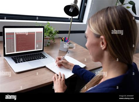 Caucasian Female Programmer Siting At Desk At Laptop With Coding On Screen Making Notes Coding