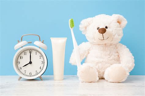 미소 사랑스러운 흰색 테디 베어 앉아서 칫솔을 들고 밝은 파란색 벽에 선반에 치약의 알람 시계와 튜브 파스텔 색상입니다 어린이