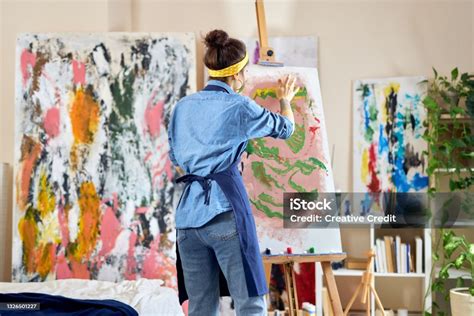 앞치마에 있는 젊은 여성 예술가의 백뷰로 이젤에 추상적인 그림을 만들고 손가락으로 캔버스에 페인트를 바르고 홈 스튜디오의 거실에 서 있습니다 25 29세에 대한 스톡 사진