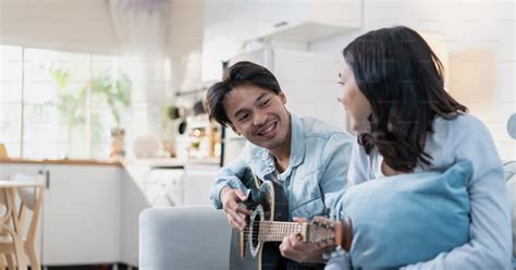 아시아의 젊은 사랑하는 커플은 집 거실에서 함께 기타를 연주합니다 매력적인 낭만적 인 새로운 결혼 소파에 앉아있는 활동적인