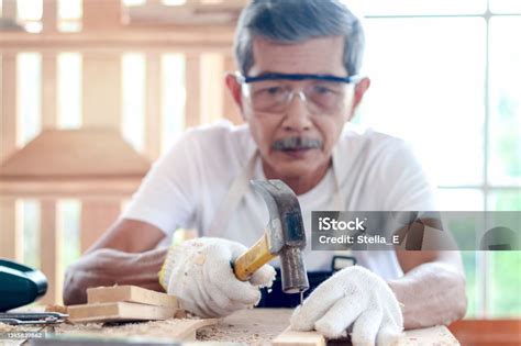 안전 안경을 쓰고 있는 노인 목수는 망치를 들고 나무 판자에 못을 박고 목공 목공 작업장에서 나무 책상에서 일하는 수석 장인이