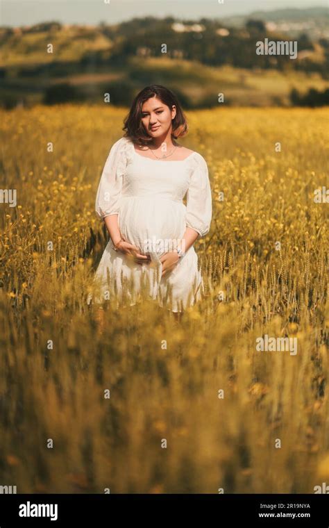 A Pregnant Woman In A White Dress Stands Elegantly In A Golden Wheat
