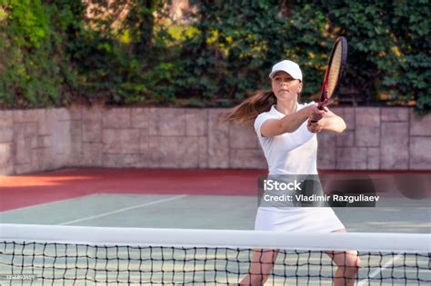 흰색 드레스와 야구 모자를 쓴 한 여성이 여름에 테니스 코트에서 잔디 테니스를 치고 라켓으로 공을 치거나 피치 스포츠 및 레크리에이션을 합니다 건강한 생활방식에 대한 스톡