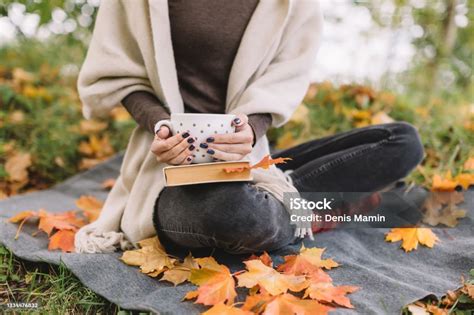 갈색 스웨터 검은 청바지 베이지 색 스카프를 입은 귀여운 소녀가 공공 공원의 격자 무늬에 앉아 책을 읽고 흰색 물방울 무늬