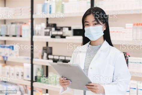 젊은 아시아 여성 약사 약사 마약 약국에서 카메라를 보면서 클립 보드를 들고 코로나 바이러스에 대한 흰색 코트와 보호 의료