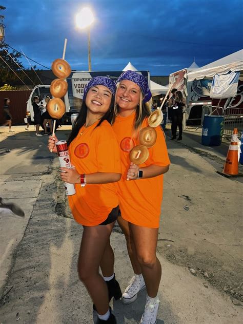 Two Women In Orange Shirts Holding Donuts And Drinking Sodas While