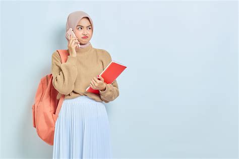 배낭으로 갈색 스웨터를 입은 젊은 아시아 무슬림 여성 학생을 괴롭 히고 휴대 전화로 이야기하고 흰색 배경에 고립 된 책을 들고