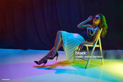 하얀 의자에 앉아 네온 불빛의 짙은 보라색 배경 위로 머리를 뒤로 젖히는 세련된 의상을 입은 젊은 여성의 가로 이미지 고급