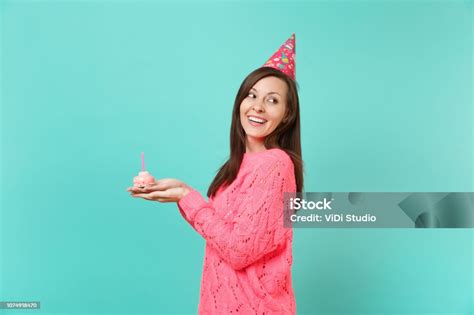 멋진에 젊은 여자의 측면 보기 핑크 스웨터 생일 모자 다시 찾고 파란색 배경 스튜디오 초상화에 고립 된 촛불 케이크를 들고 짠