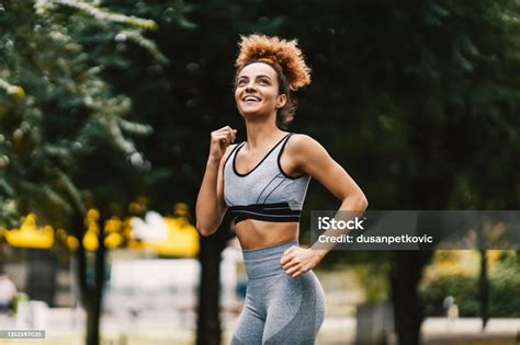 행복한 슬림 한 도시 주자가 그녀의 얼굴에 이빨미소를 지으며 도시 공원에서 달리고 있습니다 언제 어디서나 피트니스 공원에서 달리는 도시 주자 가냘픈에 대한 스톡 사진 및