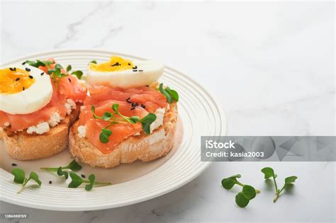 맛있는 토핑 훈제 연어 계란 허브 마이크로 그린 무 흰색 대리석 테이블 배경에 흰색 접시 위에 검은 참깨를 샌드위치 건강한 오픈 샌드위치 슈퍼 푸드 맨 위 보기입니다 연어