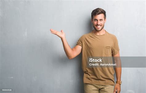 웃 고 쾌활 한 제시 하 고 카메라를 보고 손바닥으로 가리키는 회색 그런 지 벽에 잘생긴 젊은 남자 가리키기에 대한 스톡 사진
