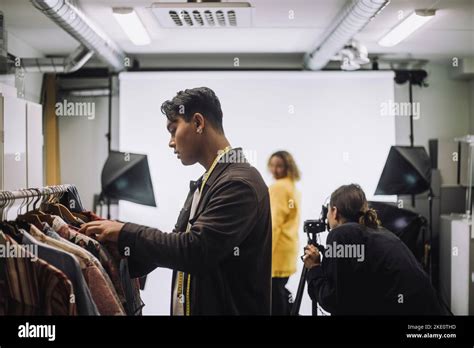 Side View Of Fashion Designer Choosing Clothes From Rack During Photo
