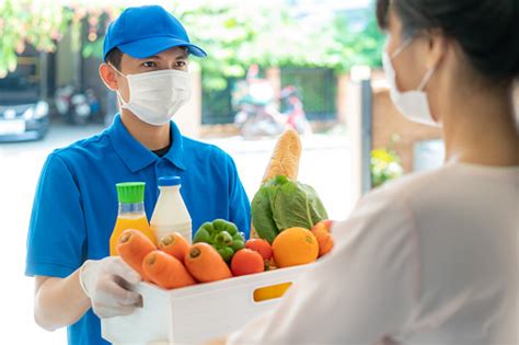 얼굴 마스크와 장갑을 착용한 아시아 여성 의상에는 집 앞에서 배달하는 남자로부터 음식 과일 채소 음료의 식료품 상자를 받습니다 2명에 대한 스톡 사진 및 기타 이미지