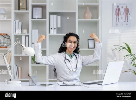 A Joyful Latino Woman Doctor With Curly Hair Raises Her Arms In A Triumphant Pose Celebrating A