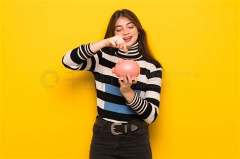 存钱罐免抠元素 黄墙上的年轻女子拿着存钱罐，很高兴，因为它装满了 素材免费下载 626像素 格式 编号69215458 千图网