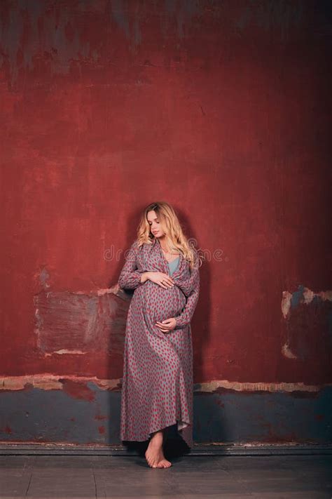 Pregnant Woman With Blond Hair In Red Dress Against Rustic Wall Stock