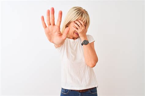 고립 된 흰색 배경 위에 서있는 캐주얼 한 티셔츠를 입은 중년 여성은 손으로 눈을 가리고 슬프고 두려움표현으로 제스처를 멈춥니다