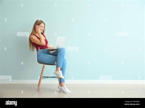 Beautiful Young Woman With Laptop Sitting On Chair Against Color Wall
