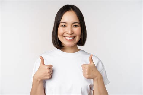 엄지손가락을 치켜들고 웃고 있는 행복한 젊은 한국 여성은 흰색 배경 위에 서 있는 Smth를 추천하는 긍정적인 피드백을
