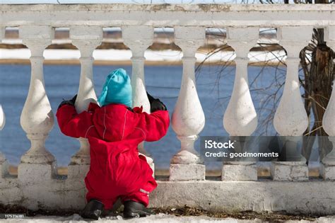 빨간 점프수트와 파란 모자를 쓰고 하얀 울타리를 쳐다보며 강을 바라보고 있는 어린 아이 해변에서 봄 화창한 날입니다 야외에서 걷고 강 전망을 즐기십시오 물에서 보고 가을에