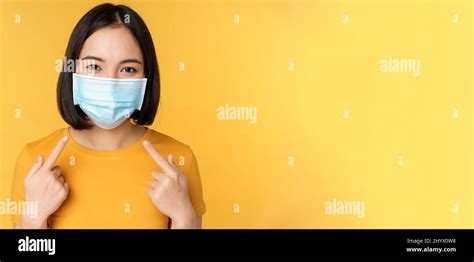 Portrait Of Smiling Asian Woman In Medical Face Mask Pointing At Her