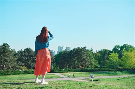 파란 블라우스와 산호 치마를 입은 빨간 머리의 십대 소녀가 공원에 서 있습니다 나무와 맑은 푸른 하늘 배경이 있는 여름