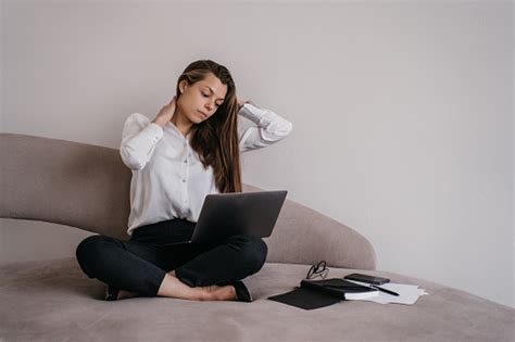 긴 머리를 가진 피곤한 갈색 머리 사업가는 열심히 일한 후 옆으로 뻗어있는 노트북을 사용하여 소파 다리에 앉아 있습니다 꽤