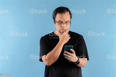 검은 색 티셔츠와 안경을 쓴 아시아 남자는 파란색 배경에 고립 된 휴대 전화를 볼 때 의심 스럽거나 무언가에 대해 생각하고