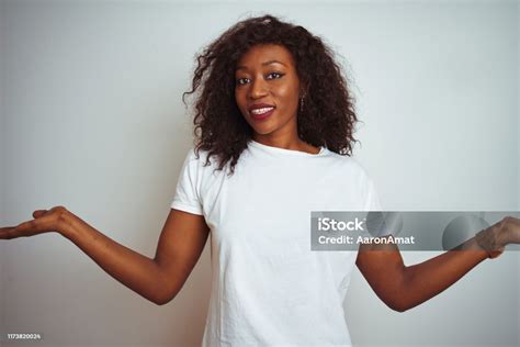 고립된 흰색 배경 위에 서있는 티셔츠를 입은 젊은 아프리카 계 미국인 여성은 양손을 열고 손바닥을 드러내며 미소 짓고 제시 및