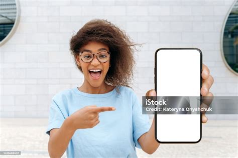 광고를위한 빈 공간 흥분한 충격에 놀란 아프리카 계 미국인 여성 야외에 서서 빈 흰색 모형 화면이있는 스마트 폰을 보여주고 손가락을 가리키고 카메라에 놀란 표정을 짓는다