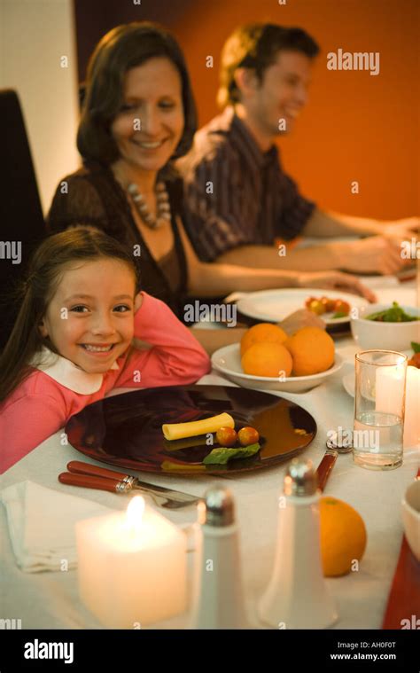 Girl Sitting At Dinner Table Smiling At Camera Adults In Background