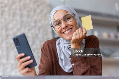 집에서 행복한 온라인 쇼핑객 여성 히잡을 쓴 무슬림 여성이 웃으며 카메라를 보고 있고 부엌에서 은행 신용 카드와 전화를 들고 있는 아랍 여성의 초상화 30 34세에 대한