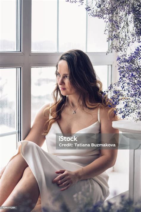 가벼운 실크 드레스를 입은 아름다운 젊은 장발 여성이 2022 년 매우 페리의 석고 필라 트렌디 한 색상으로 장식 된 방의 파노라마 창을 바라 봅니다 2022년에 대한 스톡