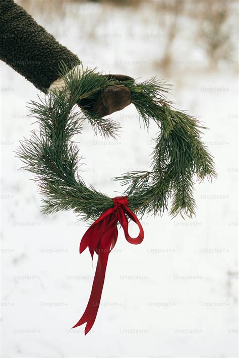 시골의 겨울 휴가 겨울 공원에서 크리스마스 화환을 가진 세련된 여자 눈 덮인 겨울 들판에서 빨간 활이 달린 크리스마스 화환을 들고 아늑한 장갑과 코트를 입은 젊은 여성