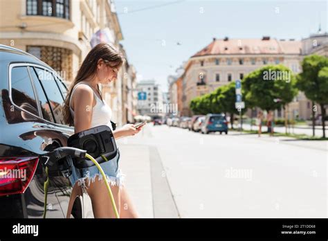 Girl In A Casual Summer Outfit Opening A Socket And Putting A Charger In A Black Electric Car