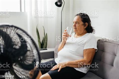 거실의 소파에 앉아있는 노인 여성은 더운 여름 날씨의 더위를 완화하기 위해 바닥 팬으로 식히고 있습니다 더운 날에 물 한 잔을