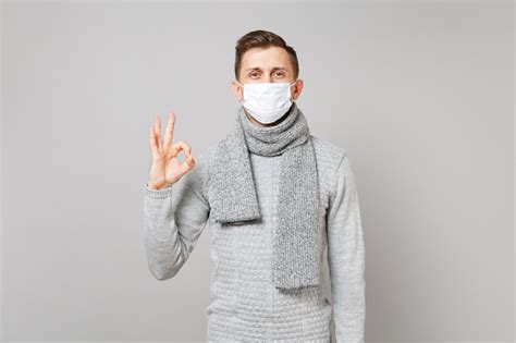 회색 스웨터에 젊은 남자 스튜디오에서 회색 배경에 고립 된 확인 제스처를 보여주는 멸 균 얼굴 마스크 스카프 건강 한 라이프