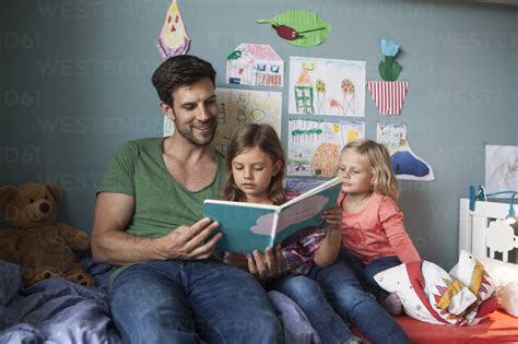 Vater und seine kleinen Töchter sitzen zusammen auf dem Bett im Kinderzimmer und lesen ein Buch