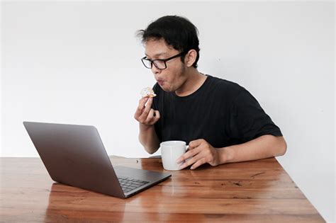 젊은 아시아 남자는 도넛을 먹고 노트북을 사용 하 여 작업 하는 동안 커피를 마시는 인도네시아 남자는 검은 셔츠를 입고 고립