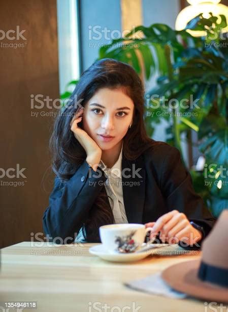 검은 색 재킷과 카페에 모자를 쓴 젊은 아시아 사업가 여성이 녹색 식물의 배경에 차 한 잔을 들고 테이블에 앉아 있습니다 25