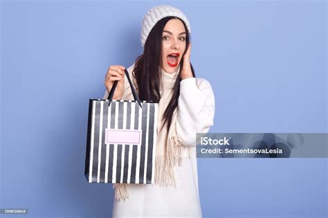남자 친구의 선물로 줄무늬 종이 가방을 들고 쾌적하고 사랑스러운 아름다운 어두운 머리 여성의 초상화를 닫고 깜짝 놀란 표정 열린 입으로 카메라를 직접 본다 가방에 대한 스톡