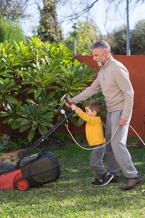Grandfather And His Grandson Mowing The Lawn In Their Home Garden Del