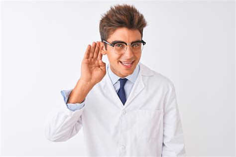 고립 된 흰색 배경 위에 안경과 코트를 입은 젊은 잘생긴 Sciencist 남자는 소문이나 험담에 귀를 듣고 귀에 손으로 미소