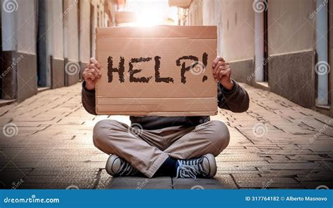 Homeless Man Sitting In The Street Holding A Cardboard Sign That Says
