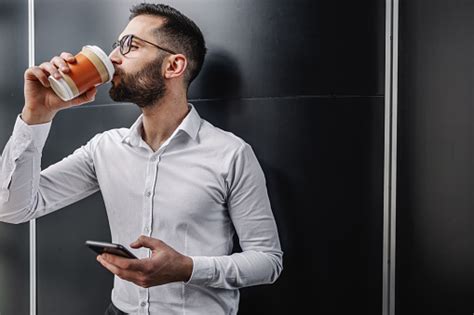 매력적인 수염 우아한 남자는 실내에 서 커피를 마시고 스마트 폰을 들고 Brand Name Online Messaging