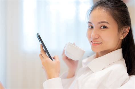 긴 머리를 하고 하얀 드레스를 입고 미소와 눈을 보내는 아시아 여성 그의 손에는 스마트폰과 커피 한 잔이 들려 있다 밝은 주변 환경 집과 같은 흰색 호텔 침실에서 25 29