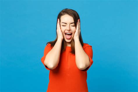 아름 다운 행복 한 젊은 여성이 빨간 오렌지 드레스를 입고 얼굴에 손을 넣어 푸른 벽 배경 스튜디오 초상화 위에 고립 승리에